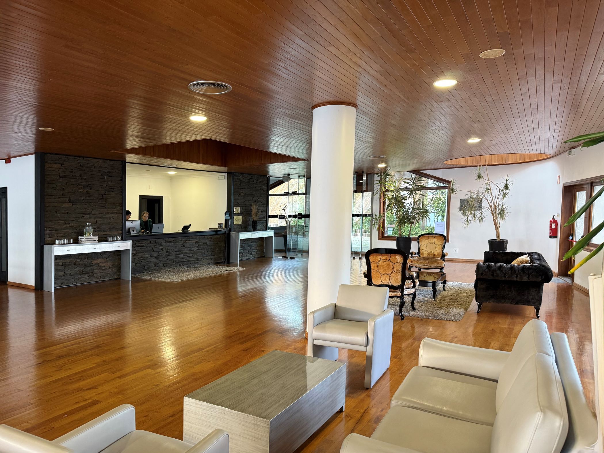 Um hall amplo com piso de madeira, balcão de recepção, área de estar e plantas em vasos.