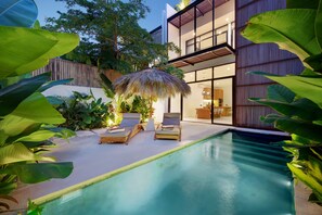 Uma casa moderna com piscina, duas espreguiçadeiras e um guarda-sol de palha.