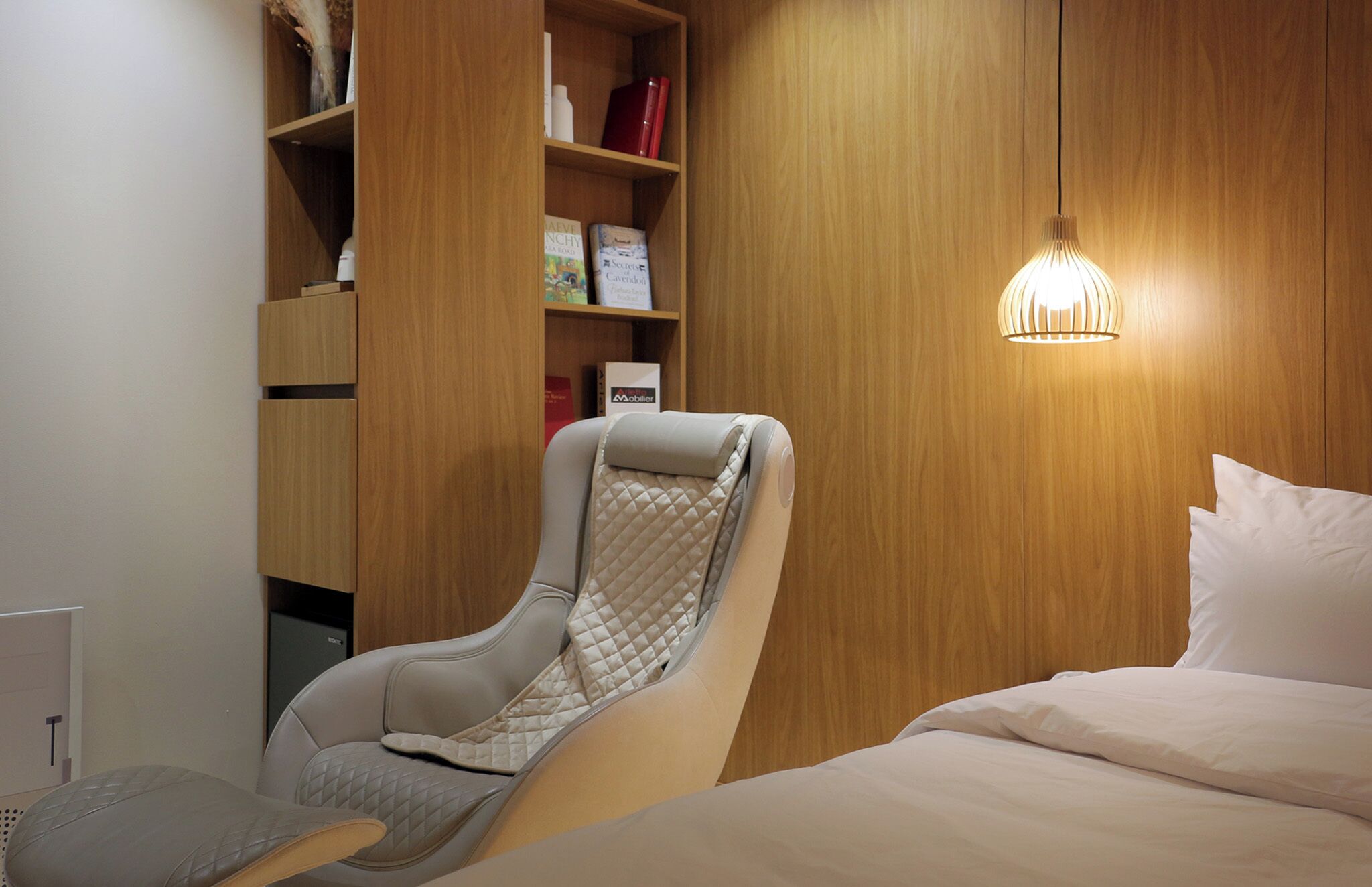 Quarto de hotel com cama, cadeira de massagem, guarda-roupa de madeira e luminária fixada na parede.