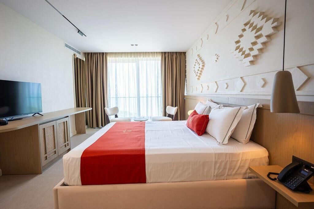 Quarto de hotel com uma cama grande, uma mesa de trabalho com televisão, telefone e janela com cortinas.