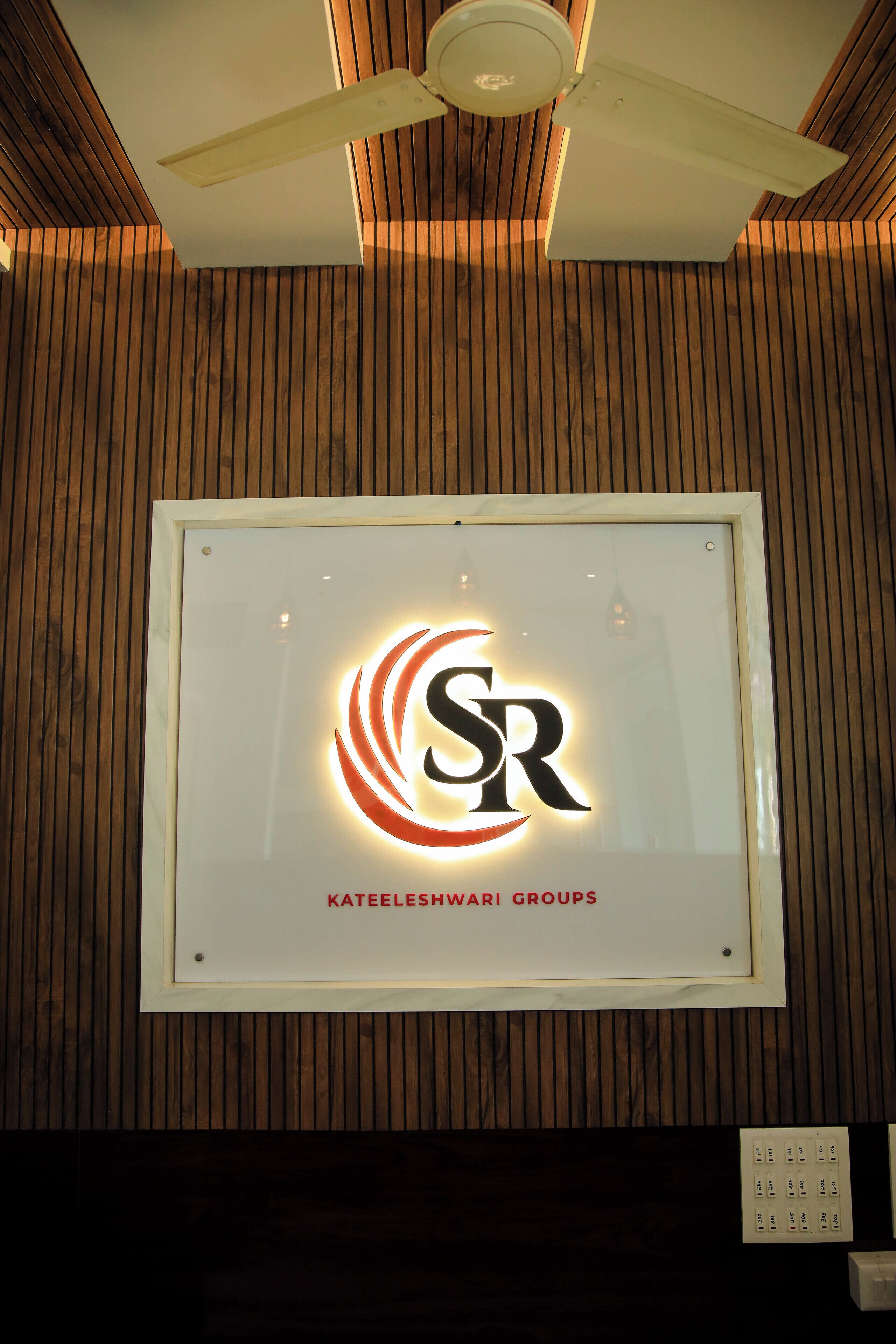 Um quadro com o logotipo e o nome de Kateeleshwari Groups numa parede de madeira.