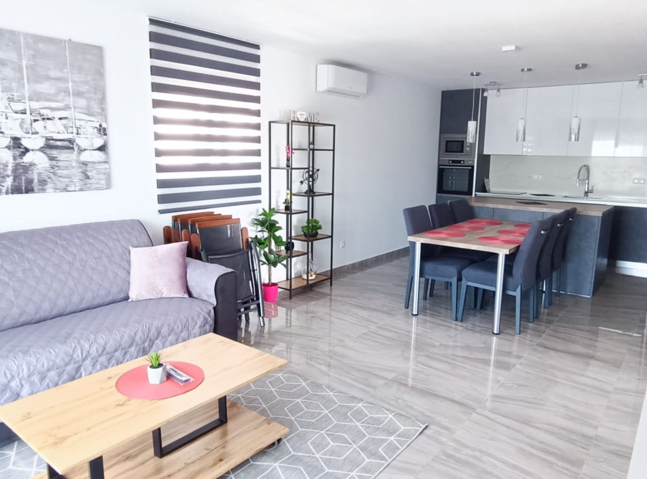 Uma sala de estar moderna com um sofá cinza, uma mesa de centro de madeira e uma área de jantar com cadeiras azuis e toalha de mesa vermelha.