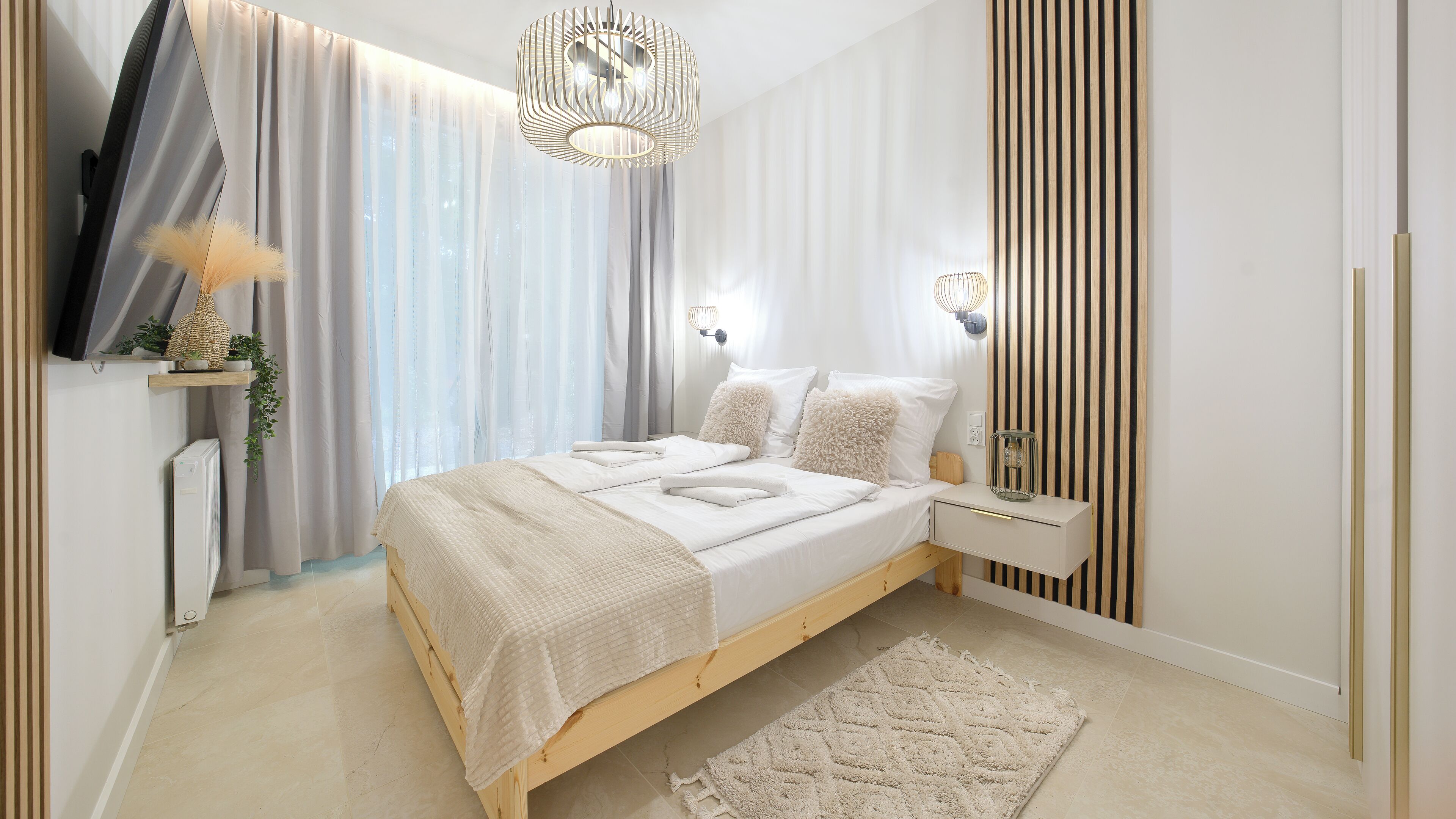 Quarto moderno com cama de madeira, roupa de cama branca, mesa de cabeceira com abajur e uma grande janela com cortinas translúcidas.