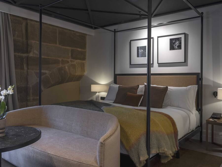 Quarto de hotel com cama, sofá, mesinha com uma planta, parede de pedra e quadros na parede.