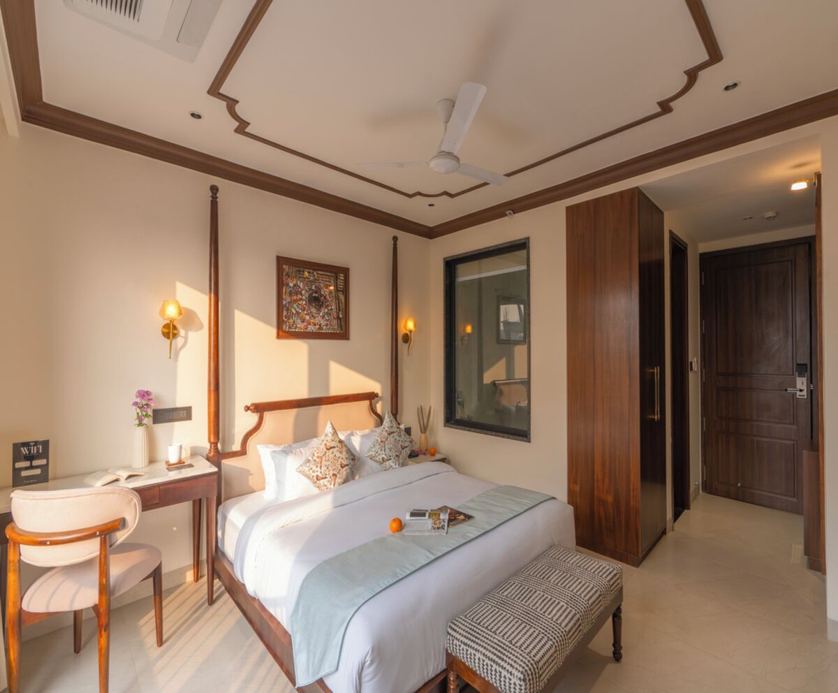 Quarto de hotel com cama grande, mesa de cabeceira, cadeira e vista para o corredor.