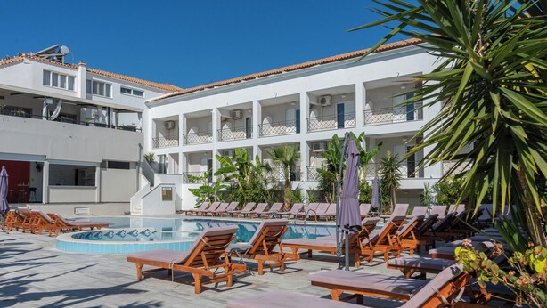 Uma área na piscina com espreguiçadeiras e guarda-sóis, céu azul límpido e um edifício com varandas.