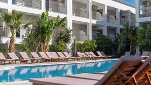Área ao lado da piscina com espreguiçadeiras e palmeiras, cercada por um edifício com várias andares e varandas.