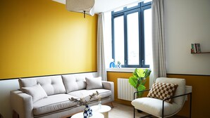 Uma sala de estar com sofá, poltrona e mesa de centro, uma janela com cortinas e um radiador.