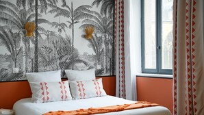 Cama com roupa de cama branca e detalhes em laranja, parede com padrão de planta tropical e janela com cortinas.