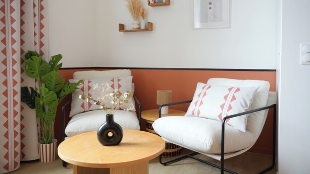 Um quarto com uma mesa de madeira redonda, duas poltronas brancas, uma planta verde grande e uma estante na parede com livros e objetos decorativos.