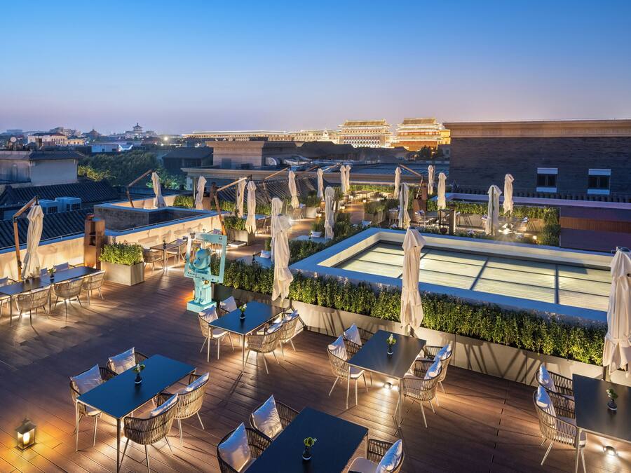 Terraço no telhado com mesas e cadeiras, uma piscina e vista para edifícios tradicionais ao fundo.