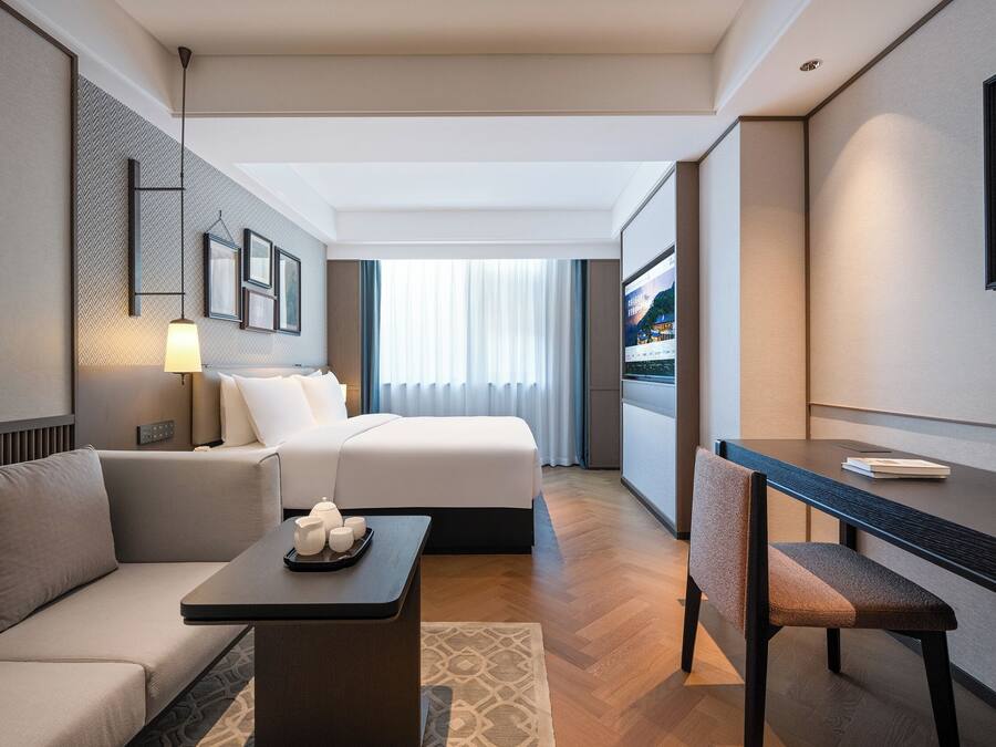 Quarto de hotel moderno com cama, sofá, escrivaninha e vista para a cidade.
