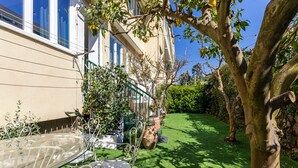 Um jardim com mesa e cadeiras, uma árvore e um edifício com janelas.