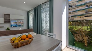 Uma sala de jantar moderna com uma mesa de madeira, uma cesta de vime com frutas e vista para uma varanda com parede de pedra e vegetação.