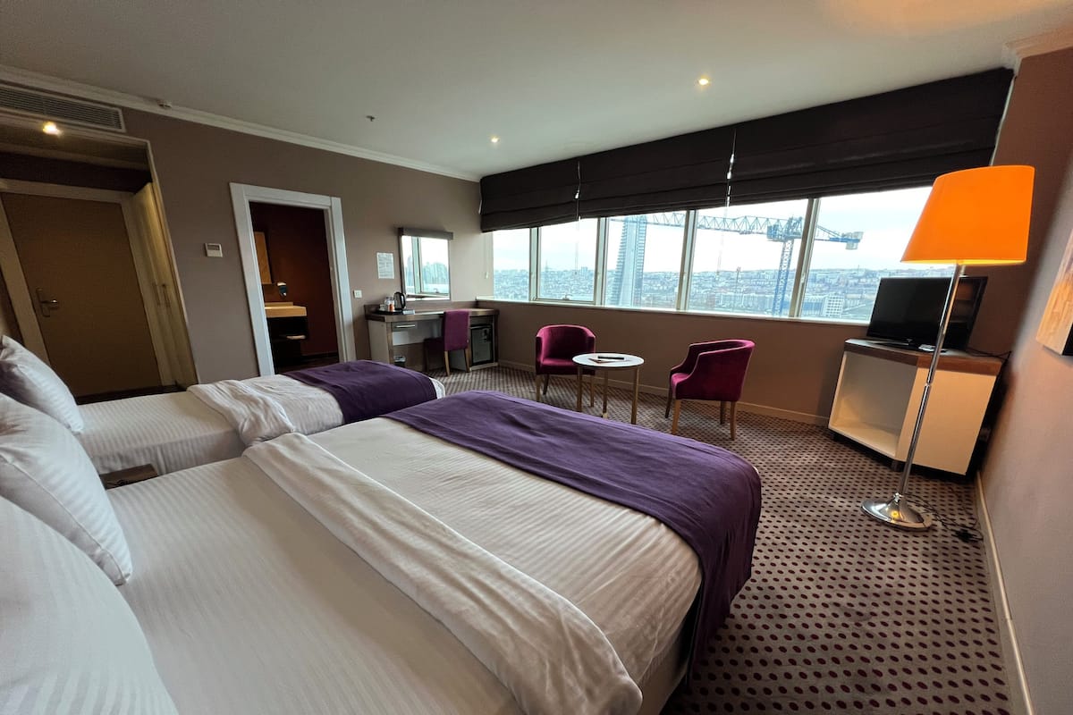 Quarto de hotel com duas camas, uma escrivaninha, uma cadeira, uma mesa pequena, um abajur e vista para a cidade.
