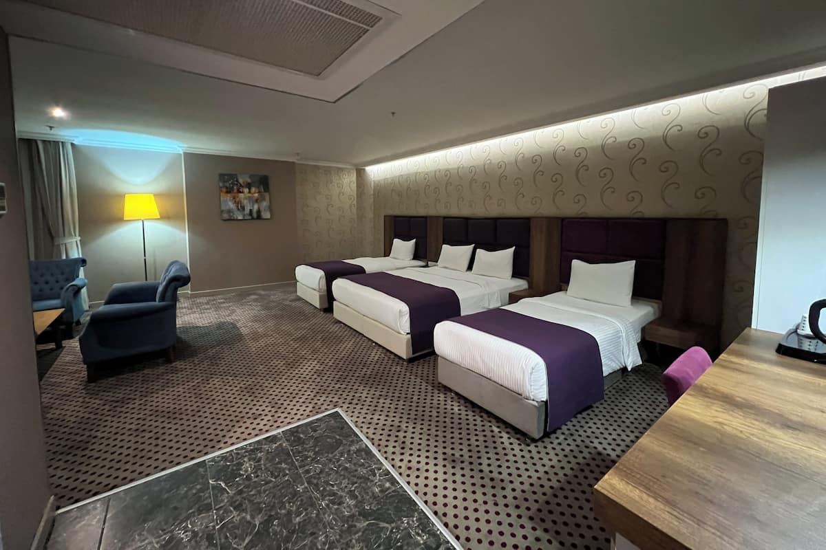Quarto de hotel com três camas, uma escrivaninha, uma cadeira, um abajur e parede estampada.