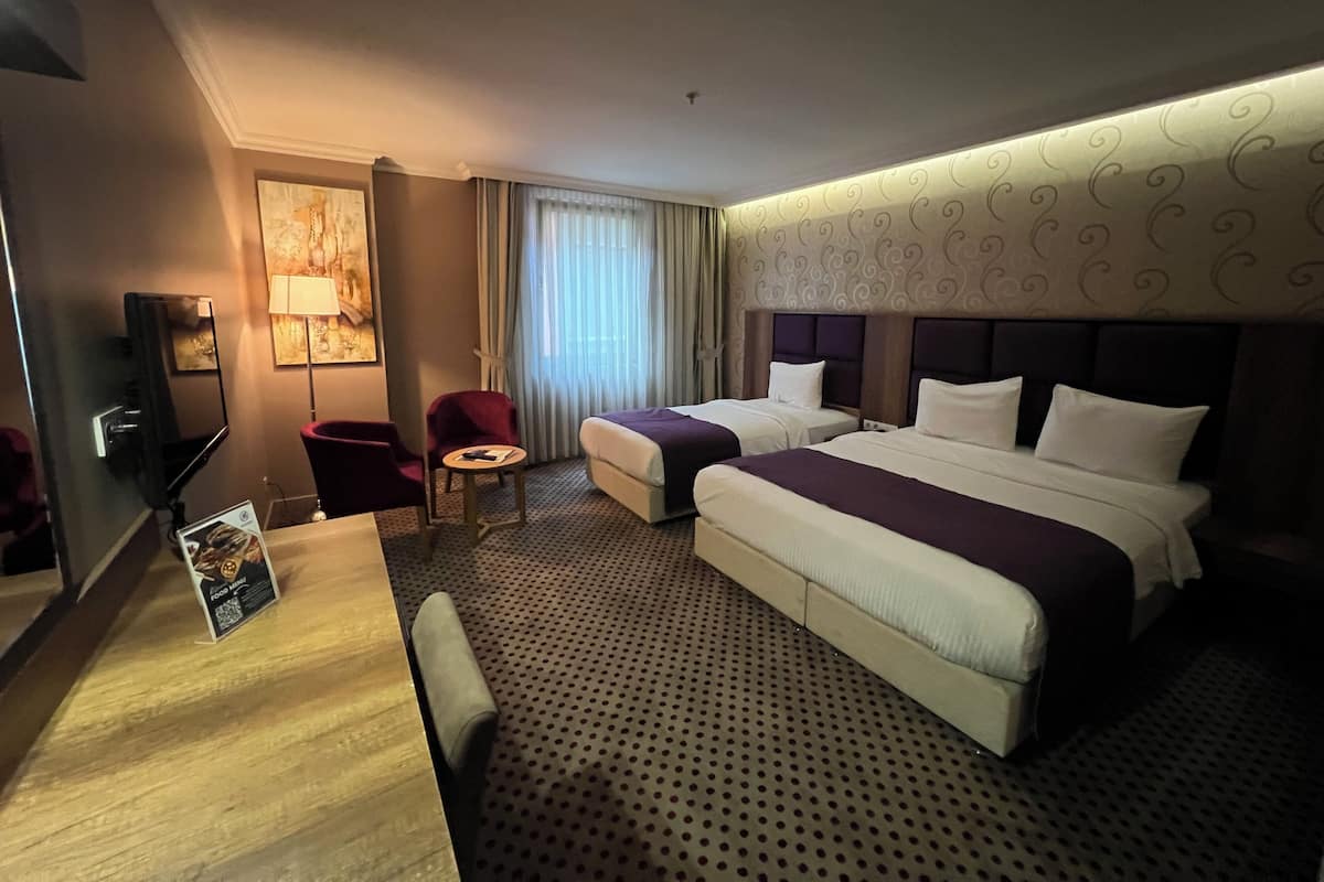 Quarto de hotel com duas camas, uma escrivaninha, uma cadeira, uma televisão e uma janela com cortinas.