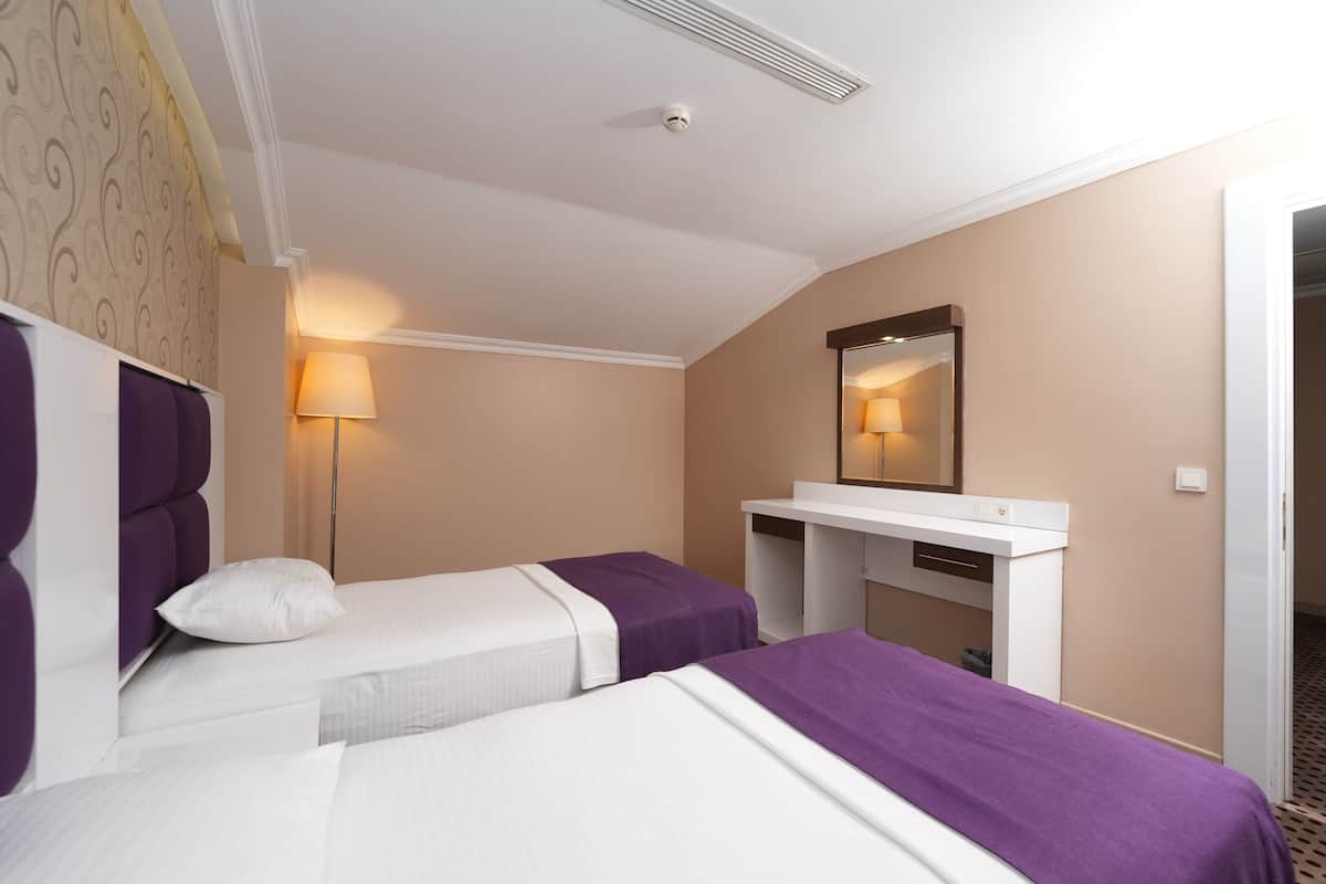 Quarto de hotel com duas camas, uma mesa de cabeceira, um abajur, um espelho e um respiradouro no teto.
