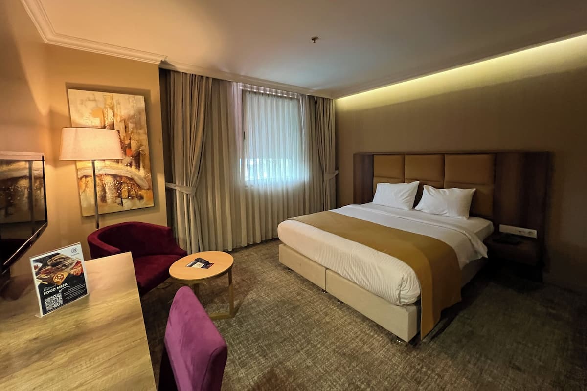Quarto de hotel com uma cama, uma secretária com cadeira, uma mesinha, um abajur e um quadro na parede.