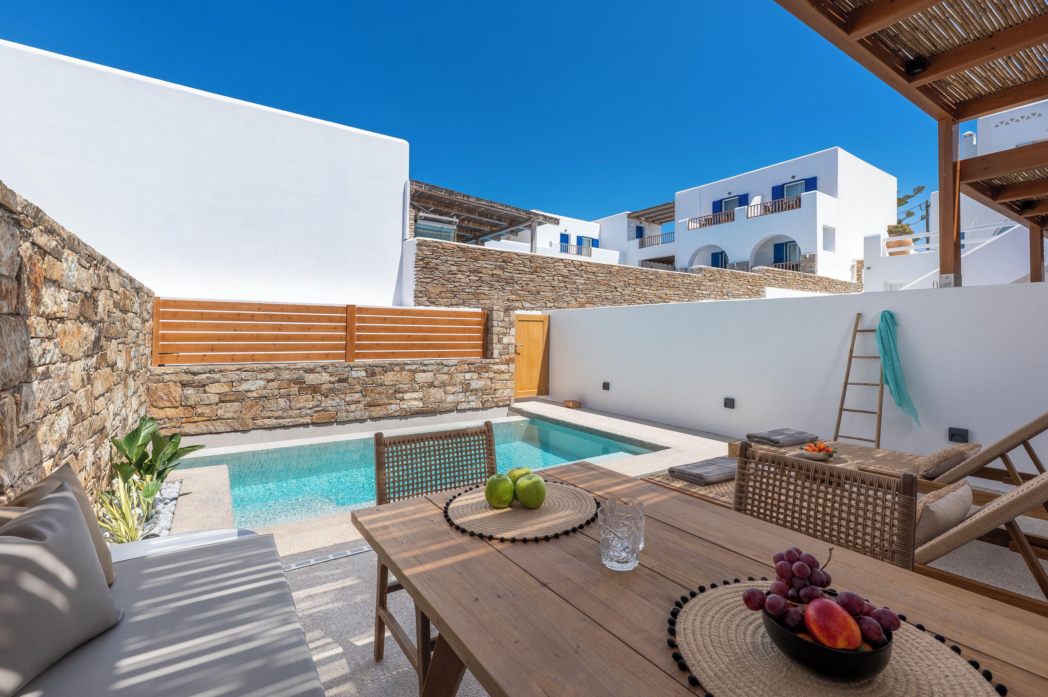 Uma área à beira da piscina com uma mesa de madeira, cadeiras de lounge e uma parede de pedra.