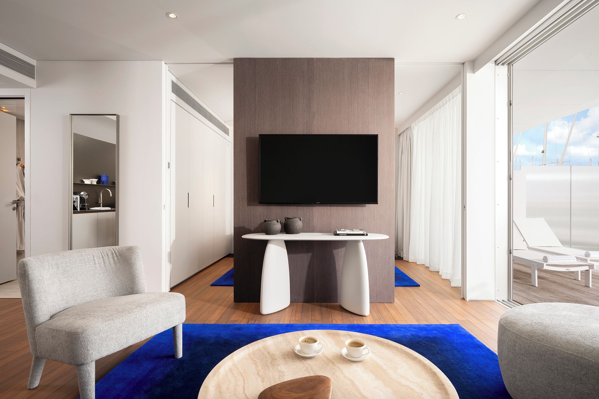 Quarto de hotel moderno com uma televisão de tela plana grande presa à parede, uma mesa de centro redonda de madeira com duas xícaras e um tapete azul.