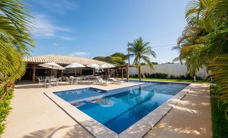 Uma piscina retangular com espreguiçadeiras e guarda-sóis, rodeada por palmeiras e um edifício com telhado de azulejos.