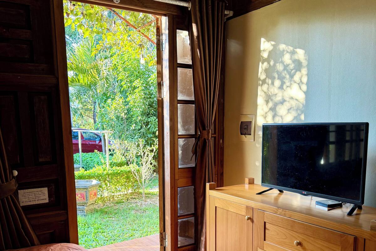 Quarto com cama, cómoda de madeira, televisão e porta aberta para um jardim.