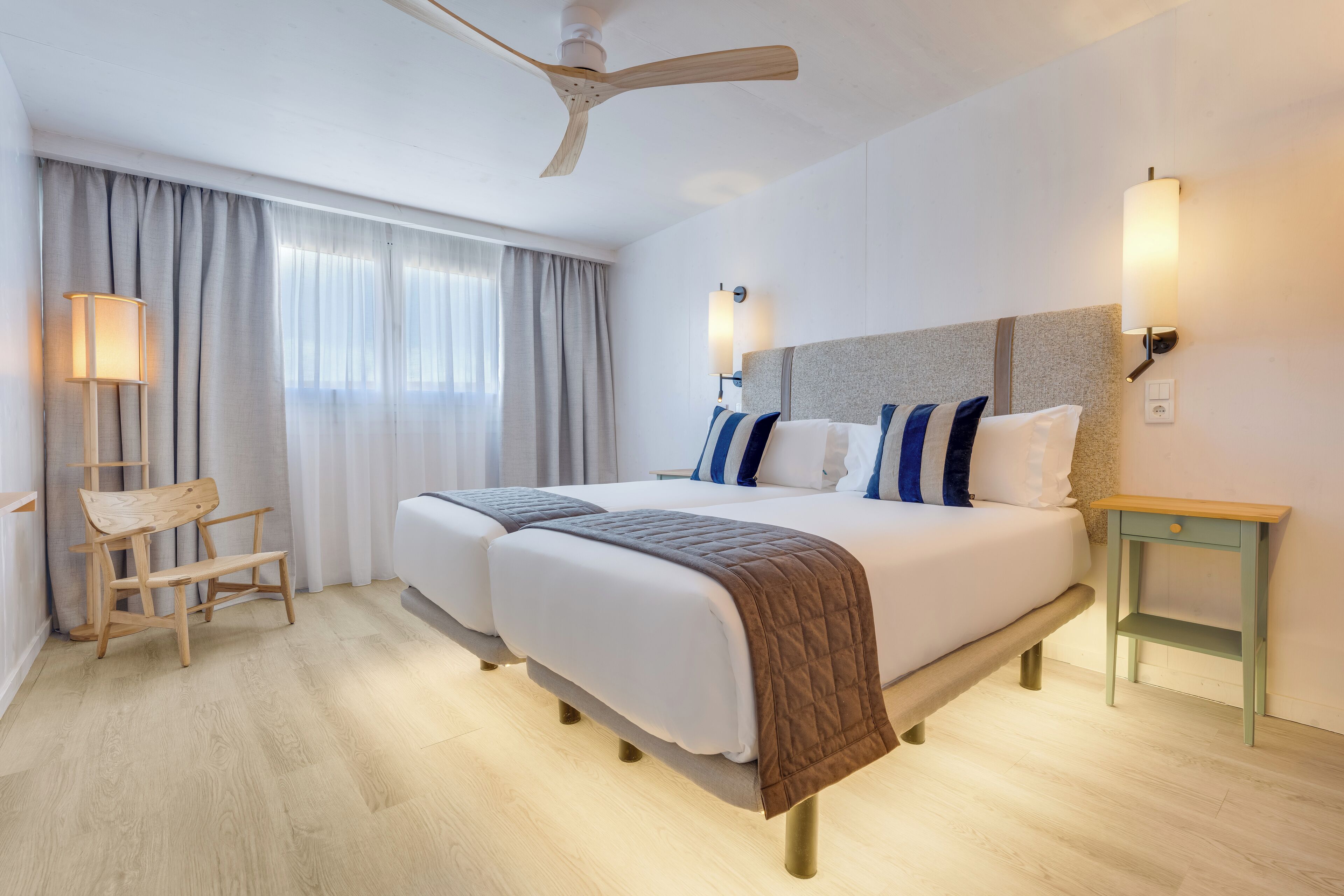 Quarto de hotel moderno com uma cama grande, duas mesas de cabeceira, um ventilador de teto e vista para o exterior através de grandes janelas.