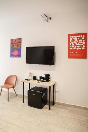 Quarto com televisão na parede, secretária com cafeteira, uma cadeira e dois quadros.