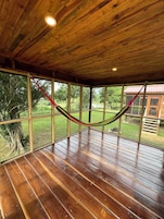 Um deck de madeira com uma rede, janelas amplas e vista para uma casa e árvores.