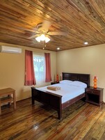 Quarto com painéis de madeira, uma cama, ventilador de teto, ar condicionado e janela com cortinas.