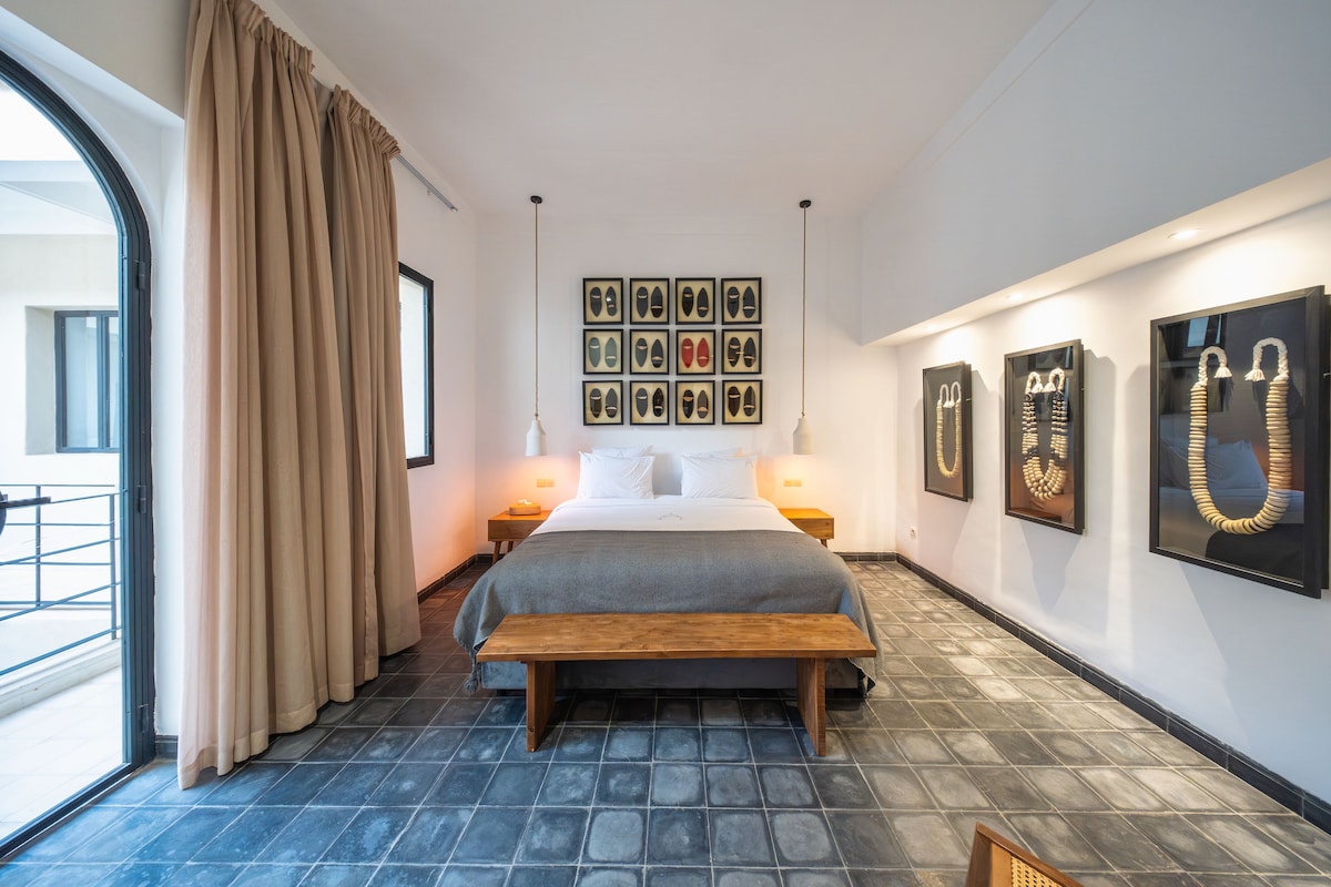 Quarto de hotel com cama, mesas de cabeceira, um banco, uma janela grande com cortinas e obras de arte penduradas na parede.