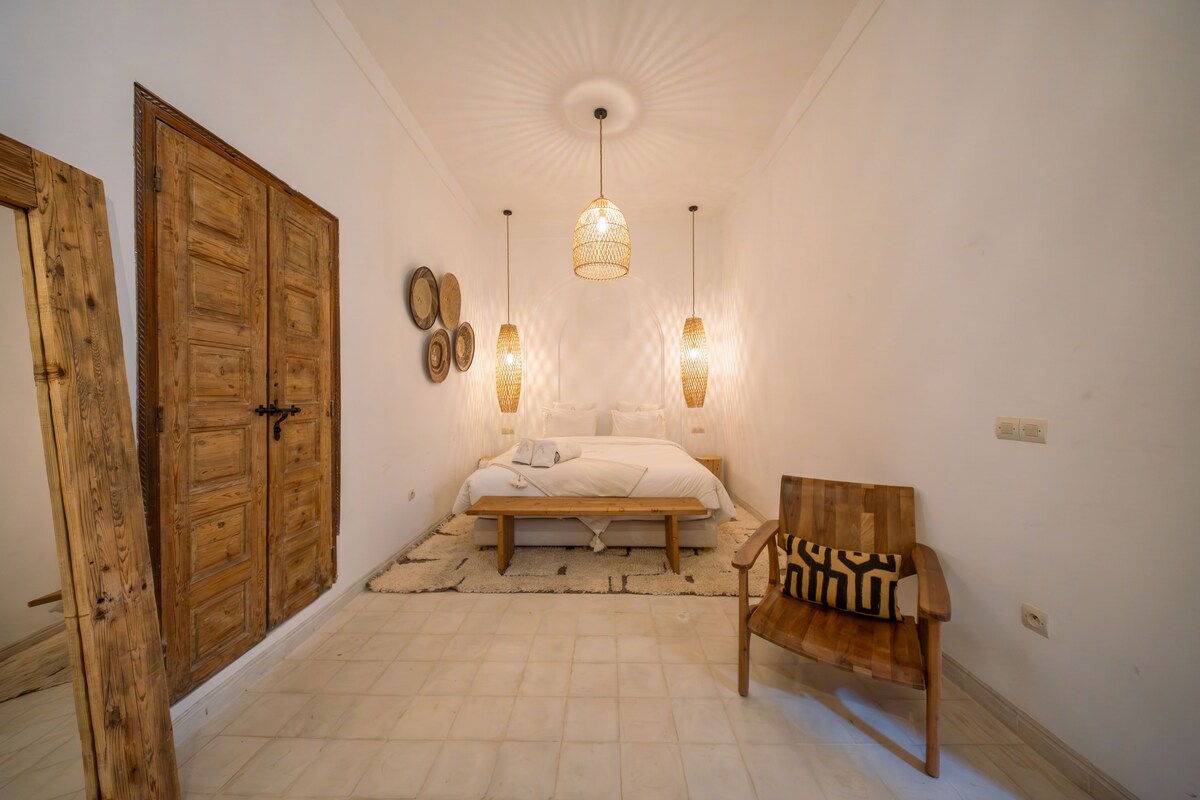 Quarto com cama, um banco de madeira e uma cadeira. Há duas portas de madeira e um teto com luminária.