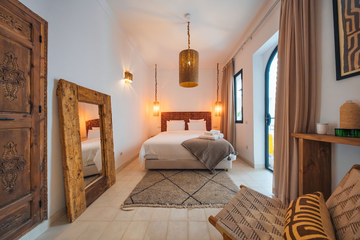 Quarto de hotel com cama grande, armário de madeira, luminária pendente e vista para o exterior.