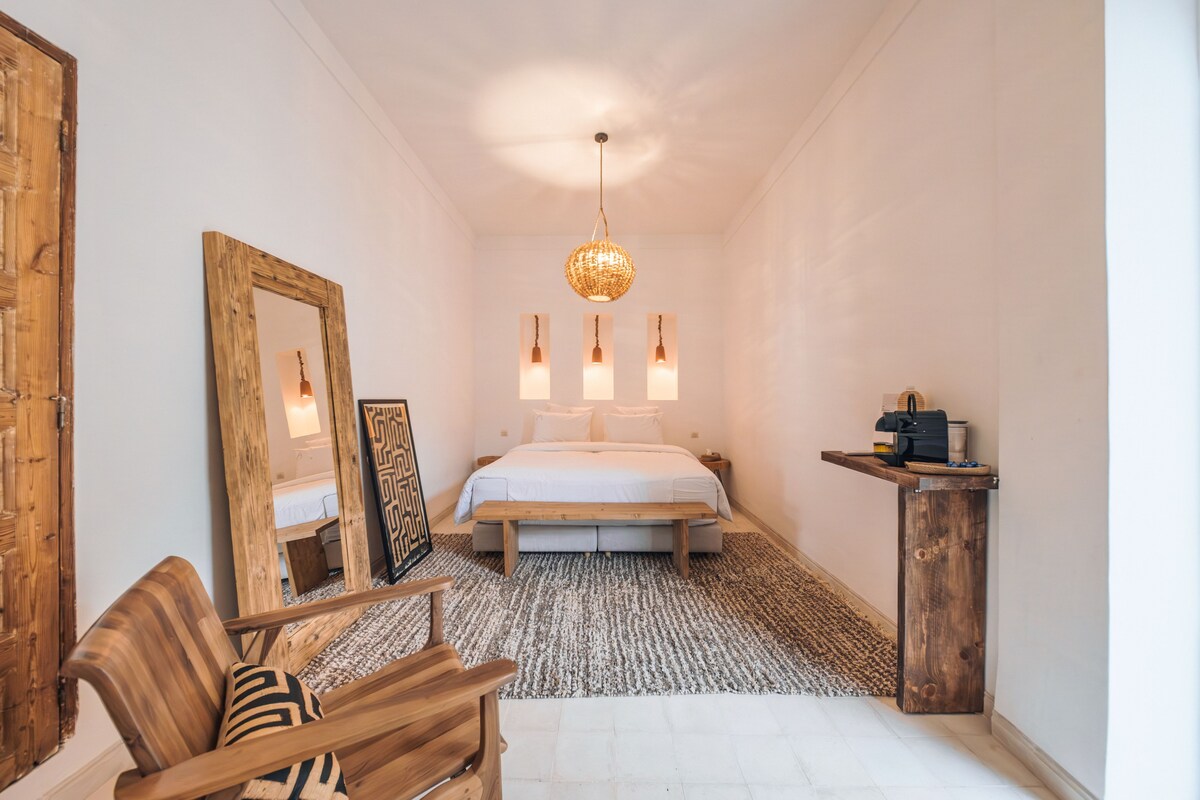 Quarto com cama, cadeira de madeira, espelho, mesa de cabeceira e luminária pendente.