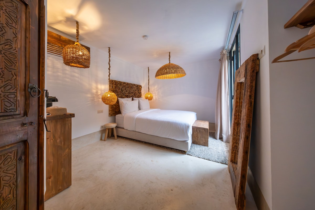 Quarto com cama, mesas de cabeceira, cabeceira de madeira e luminárias de vime suspensas.