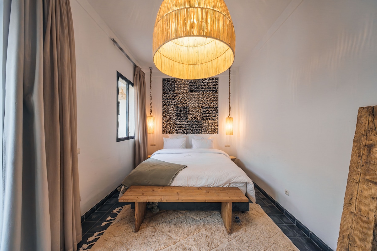 Quarto com cama, um banquinho, luminária pendente e uma obra de arte com textura na parede.