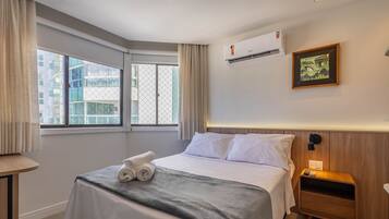 Quarto de hotel com cama, mesa de cabeceira, ar condicionado, janela com cortinas e um quadro na parede.
