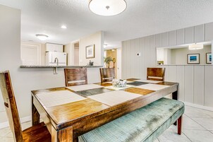 Uma área de refeições com uma mesa de madeira, cadeiras e um banco. Uma cozinha é visível ao fundo.