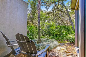 Uma cadeira Adirondack de madeira num jardim com um caminho de pedra, rodeado por vegetação exuberante e árvores.