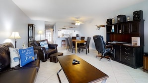 Uma sala de estar espaçosa com um sofá de couro, uma mesa de centro de madeira, e uma área de jantar com mesa e cadeiras de madeira. Há uma cadeira de escritório preta e um armário com alto-falantes.