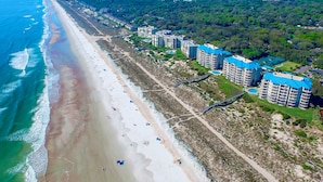 Vista aérea de uma área costeira com praia, edifícios à beira-mar e uma área arborizada.