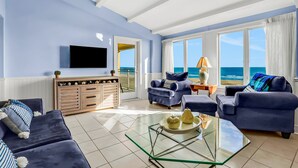 Uma sala de estar com uma grande janela com vista para o mar, uma TV de tela plana na parede e uma mesa de centro de vidro com objetos decorativos.
