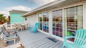 Um deck coberto ao ar livre com cadeiras, uma mesa, janelas amplas e um edifício azul-turquesa.