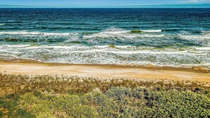 Uma praia com ondas quebrando na costa, céu claro e vegetação em primeiro plano.