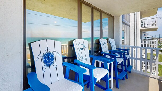 Uma varanda com cadeiras Adirondack azuis e brancas, um corrimão branco e vista para o oceano e edifícios.