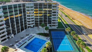 Um hotel com vários andares, piscina no terraço, quadra de tênis e vista para a praia e o oceano.