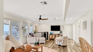 Uma sala de estar ampla com sofá, poltrona e mesa de centro. Um ventilador de teto e uma TV estão fixados na parede.