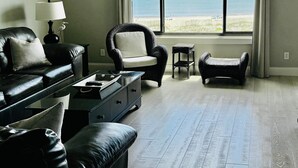 Sala de estar com vista para a praia, um sofá de couro preto, uma poltrona branca e uma pequena mesa.
