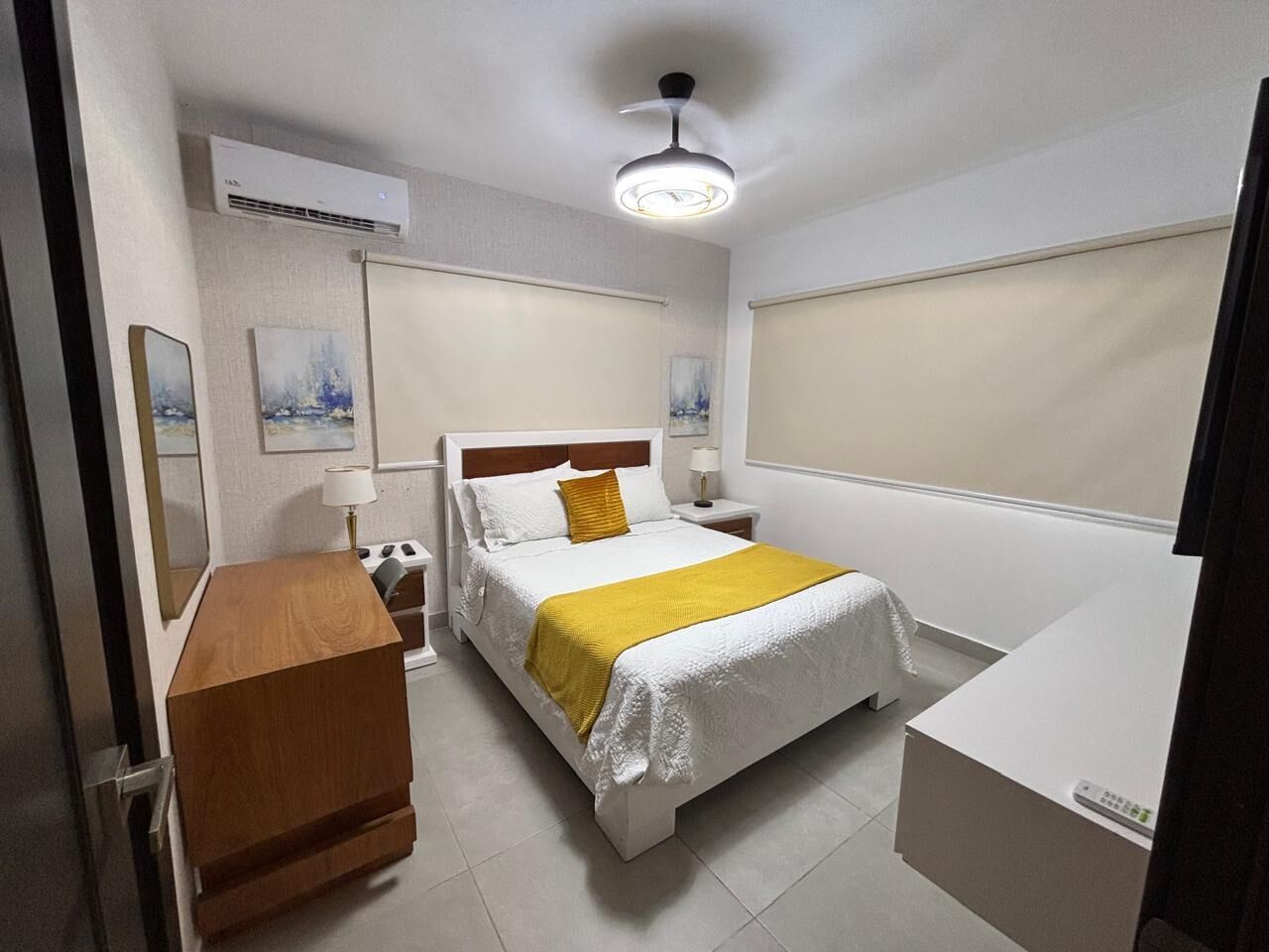 Quarto com cama, mesas de cabeceira, cômoda de madeira e aparelho de ar condicionado.